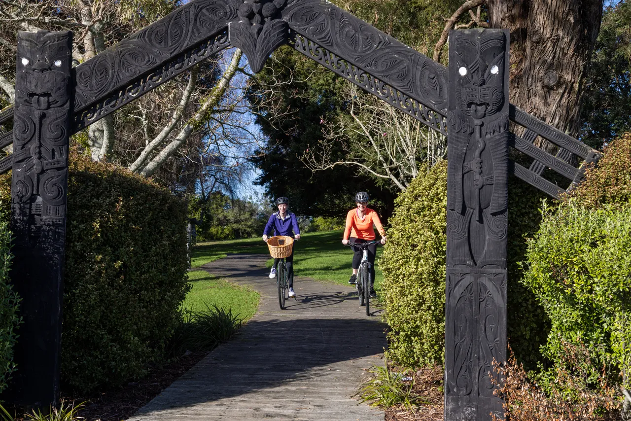 Two women cycle under the waharoa at Gate Pā Reserve